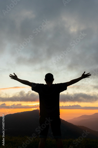 silhouette of young man standing on top of mountain peak with his hands ...