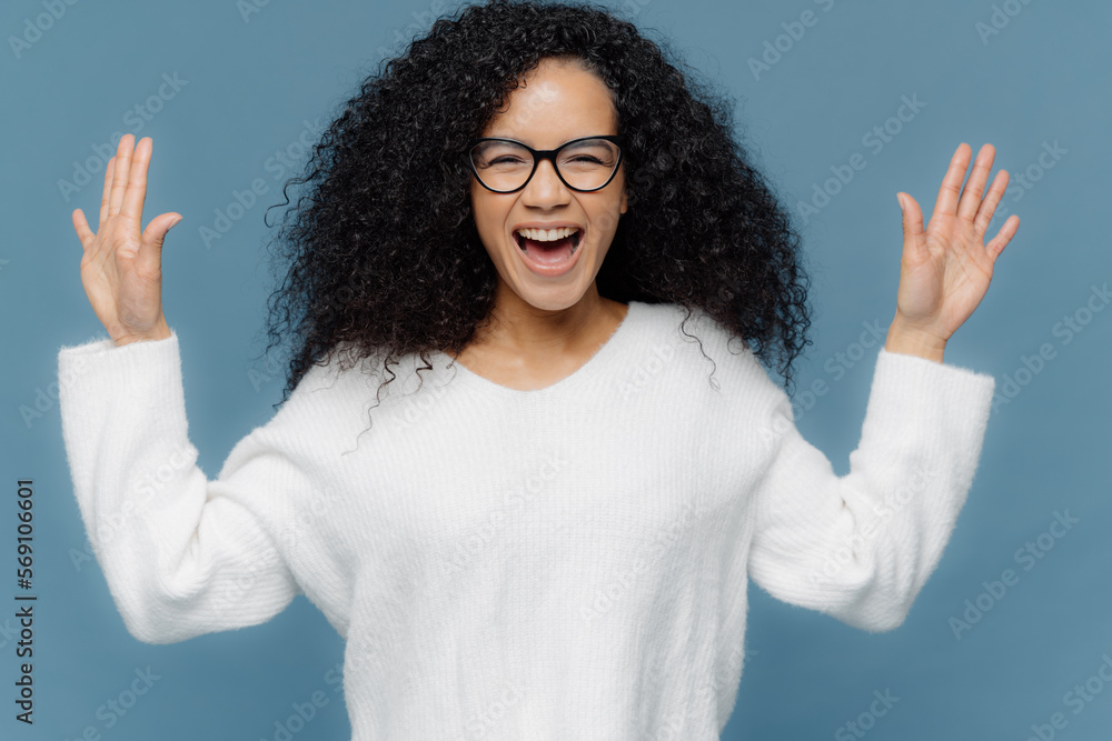 Overjoyed mixed race woman with curly hair, raises hands, exclaims from ...