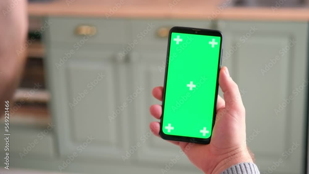 Young man sitting at home holding smartphone green mock-up screen in ...