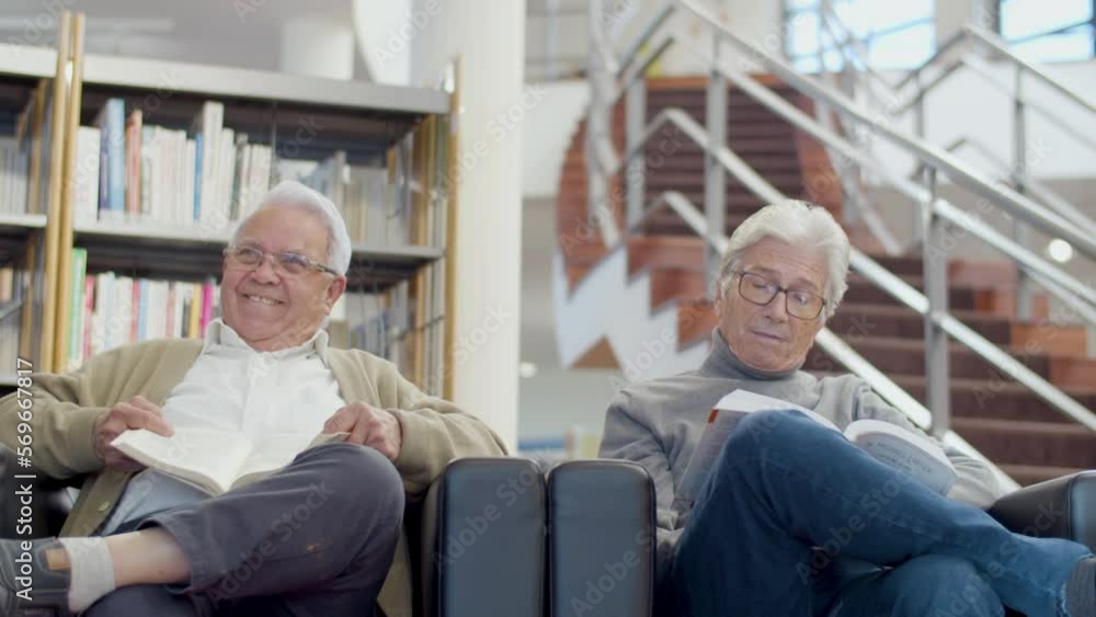 Cheerful senior friends sitting in armchairs in library. Front view of ...