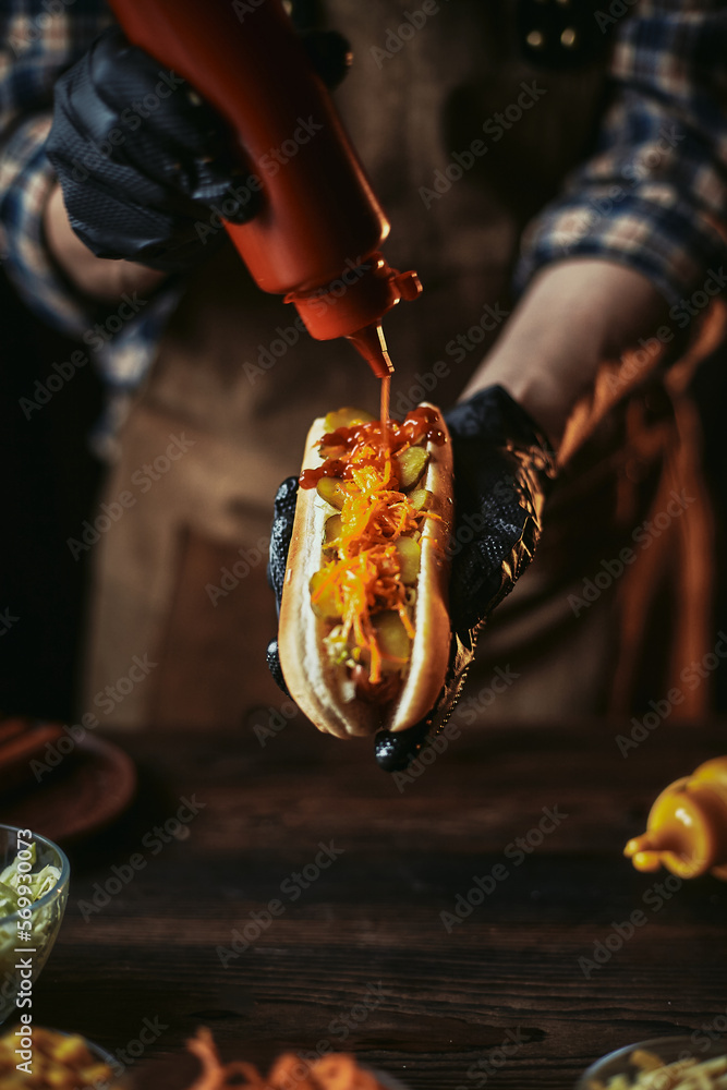 A guy in a leather apron is preparing a hot dog. The chef in gloves ...