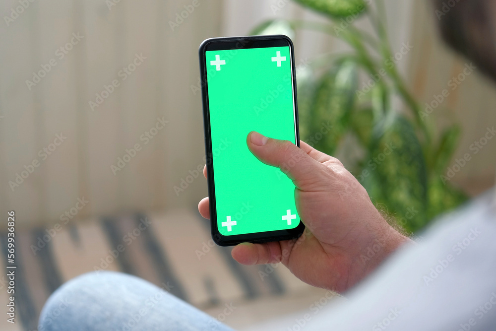 Young man sitting at home holding smartphone green mock-up screen in ...