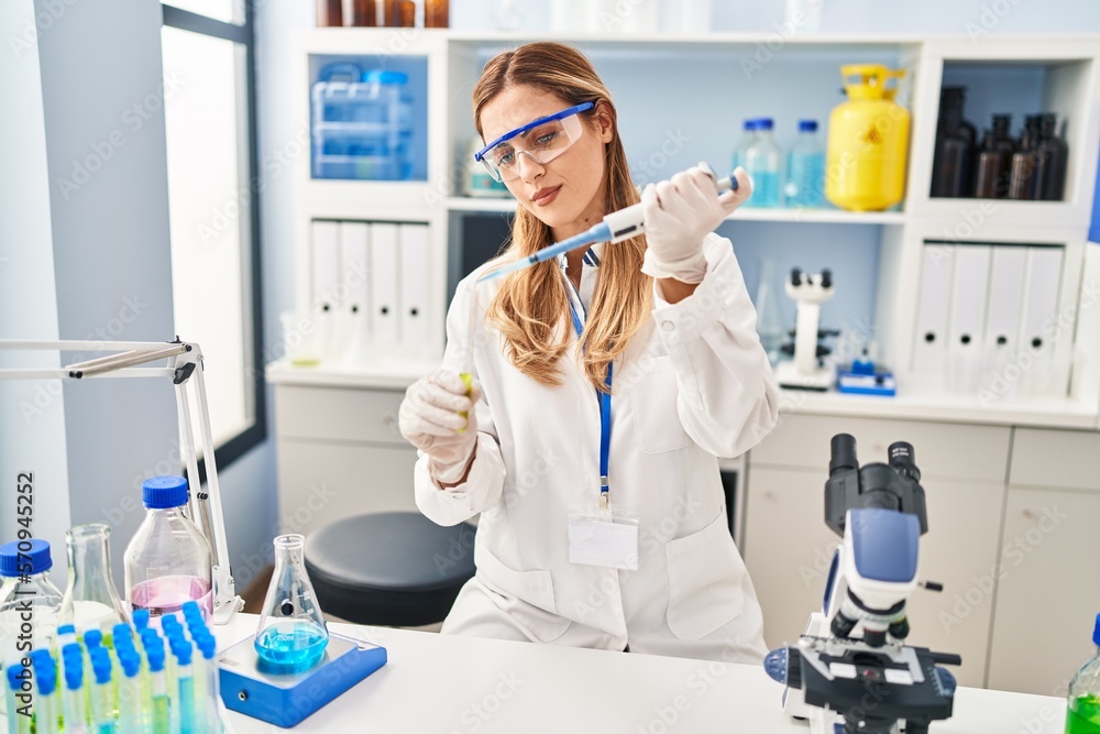 Young blonde woman wearing scientist uniform pouring liquid using ...
