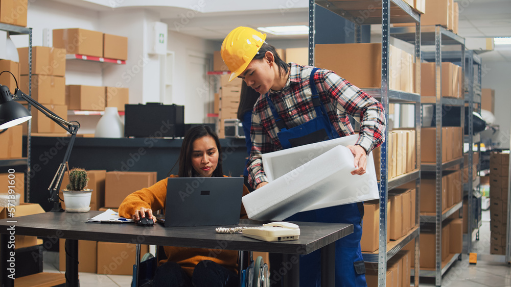 Female wheelchair user checking logistics on laptop in warehouse space ...