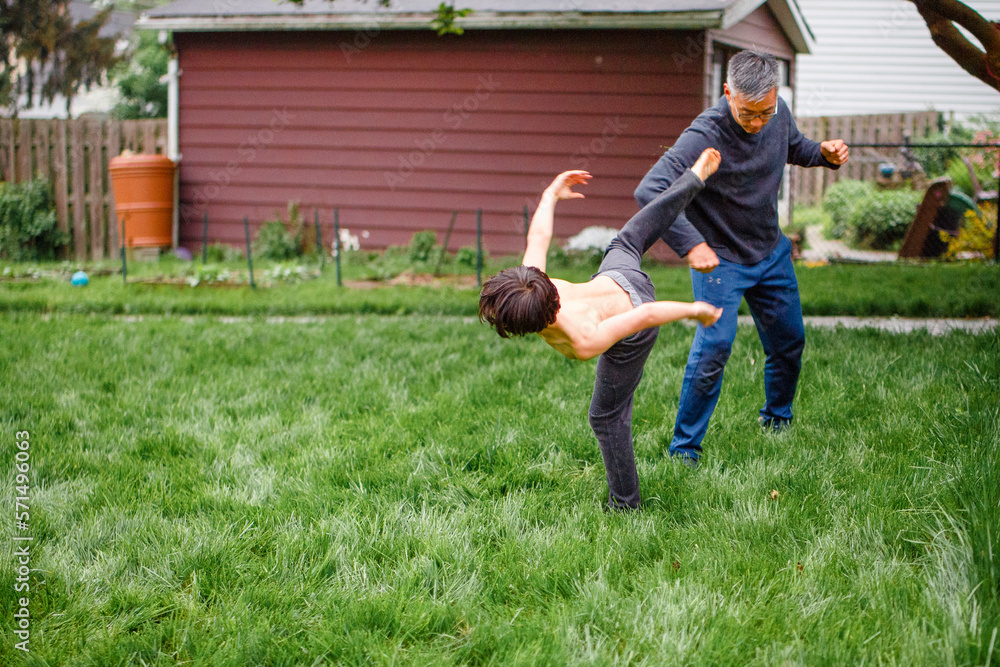 A shirtless tween boy roughhouses with father in the yard in spring ...