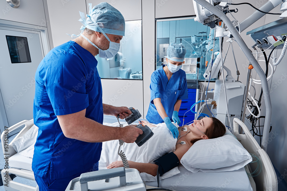 Doctor holding defibrillator on unconscious female patient's chest in intensive care unit during ...