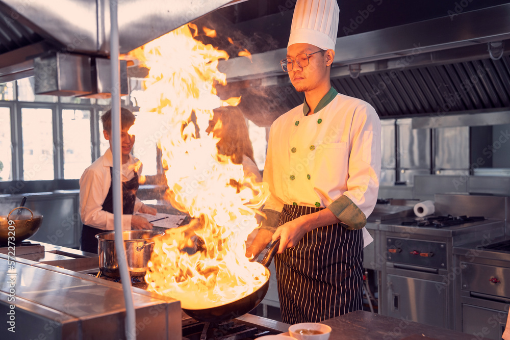 Asian male chef in uniform in chef's hat standing stir fry vegetables ...
