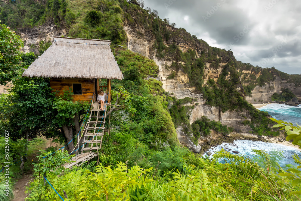 Woman sitting outside thatched roof hut on coast with cliffs, Nusa ...
