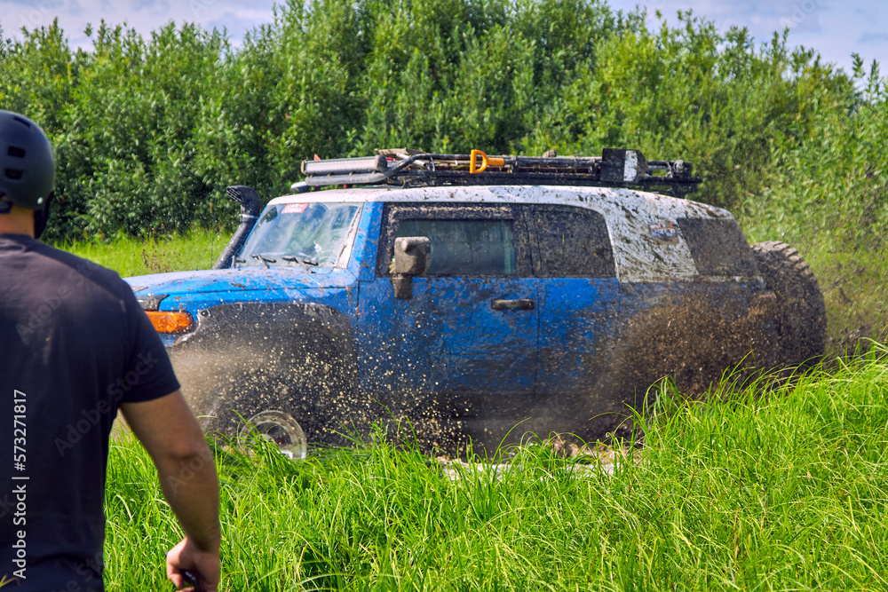 Athlete in a helmet watches as a blue off-road car splashing mud passes ...