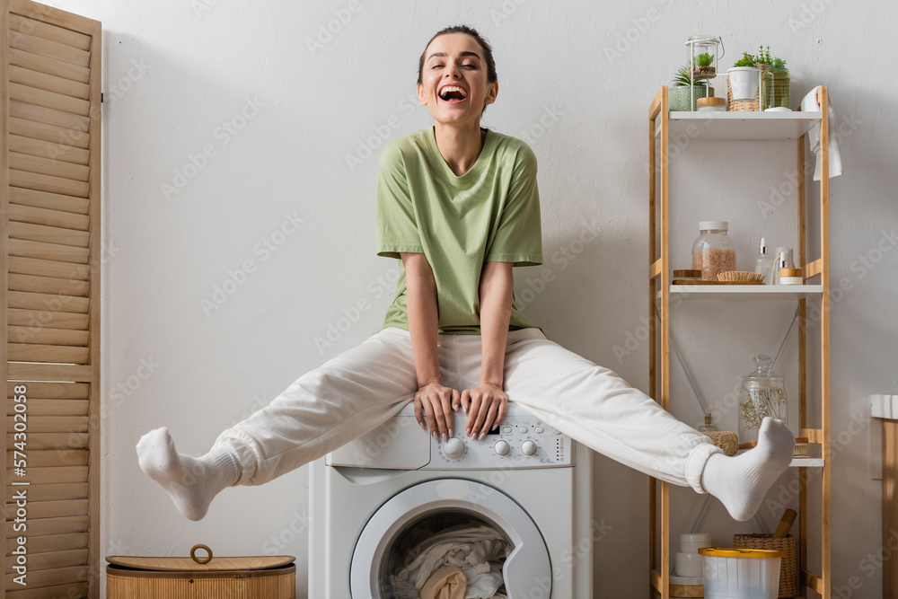 Excited woman sitting on washing machine with clothes at home. Stock ...