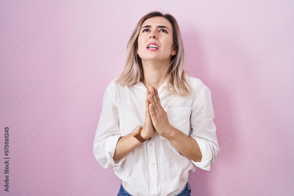 Young beautiful woman standing over pink background begging and praying ...