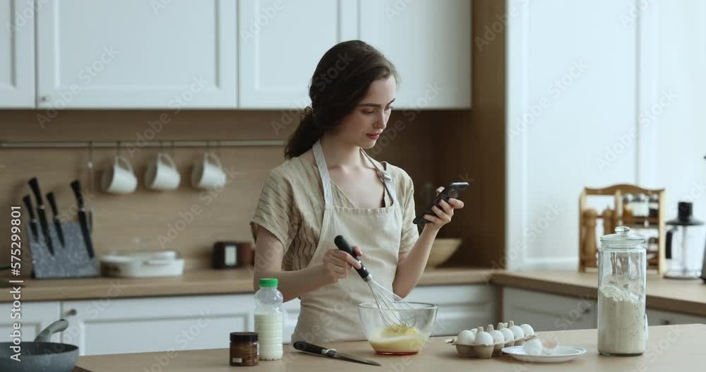 Young woman in apron cook alone in the kitchen, holds cell phone, read ...