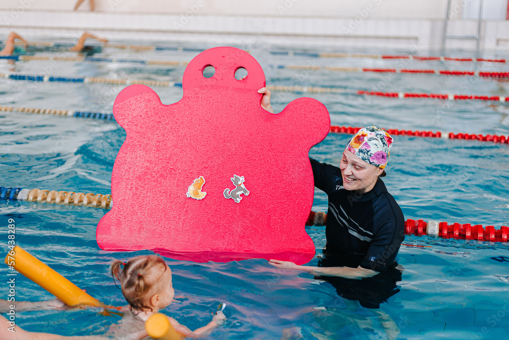 Little girl learning to swim in indoor pool with pool board and trainer ...