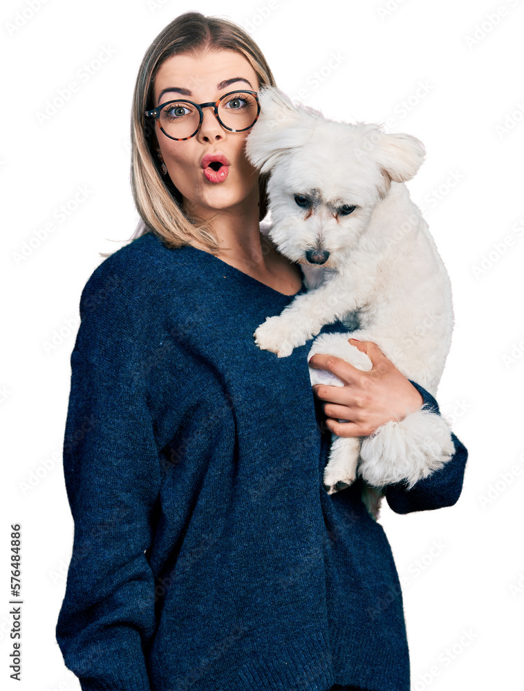 Young blonde woman hugging cute dog scared and amazed with open mouth ...