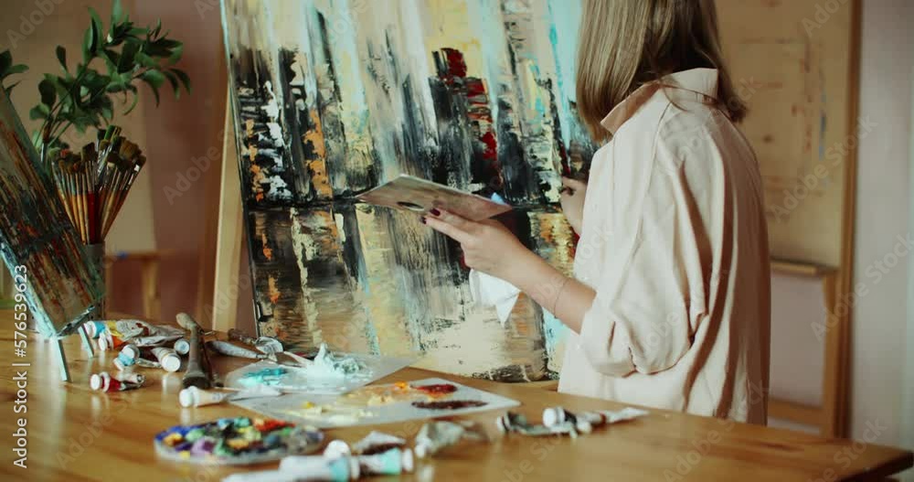 Talented innovative blondy woman artist paints oil with spatula on canvas in art workshop. She ...