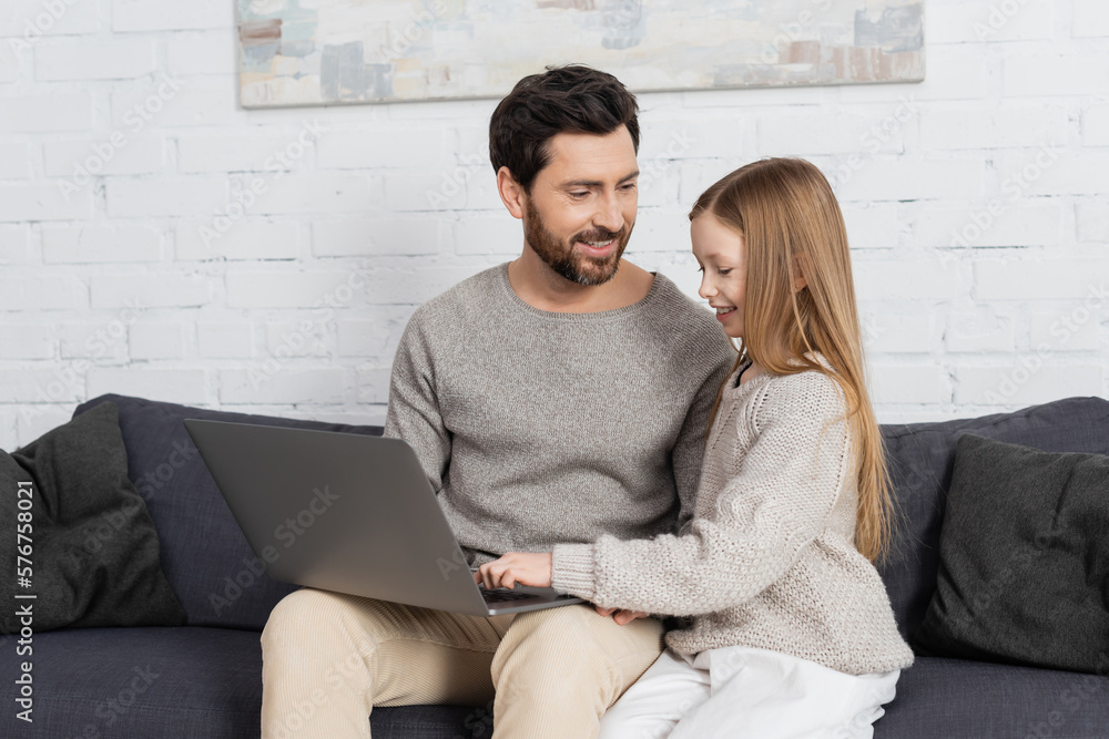 preteen girl using laptop near smiling bearded dad sitting on sofa at ...