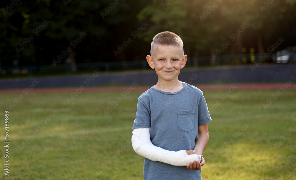 Smiling little boy with broken hand looks at camera outdoors on sports ...