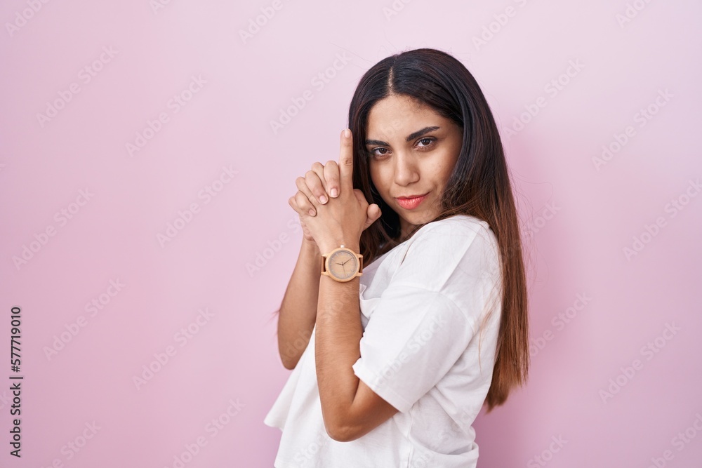 Young arab woman standing over pink background holding symbolic gun ...