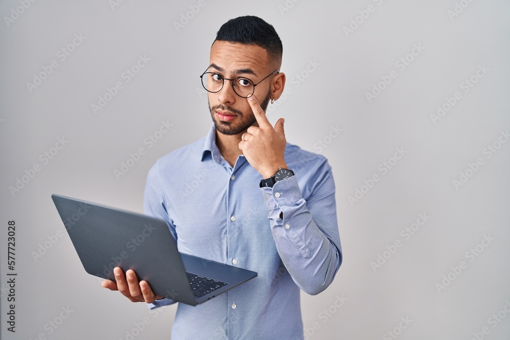 Young hispanic man working using computer laptop pointing to the eye ...