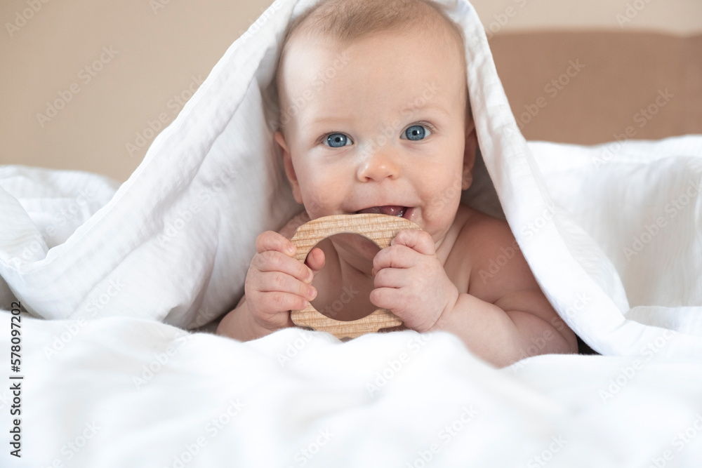 Cute happy baby with wooden teether in hands laying on tummy in bed ...