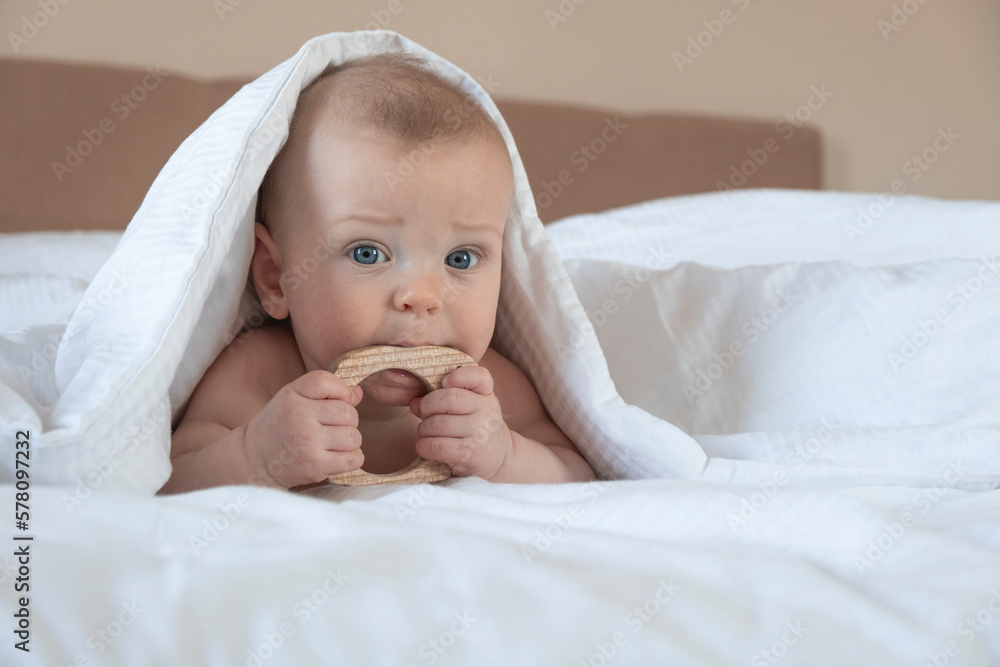 Cute happy baby with wooden teether in hands laying on tummy in bed ...