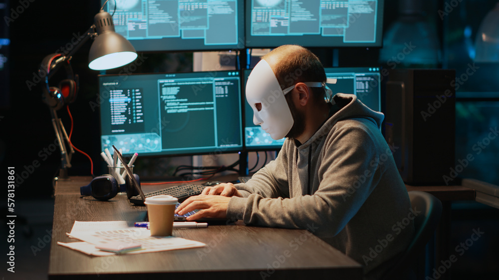 Masked IT thief hacking computer system late at night, working on laptop and multiple monitors ...