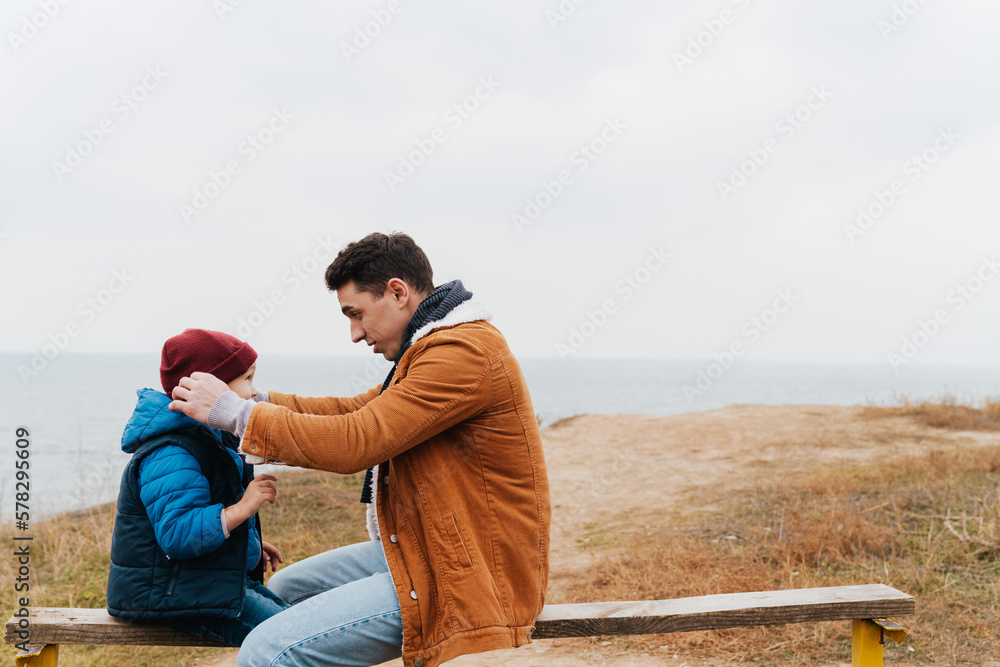 Father putting hat on his son's head while sitting together on bench at ...