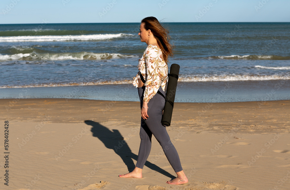 pretty woman walking barefoot on the beach with mat slung over her ...