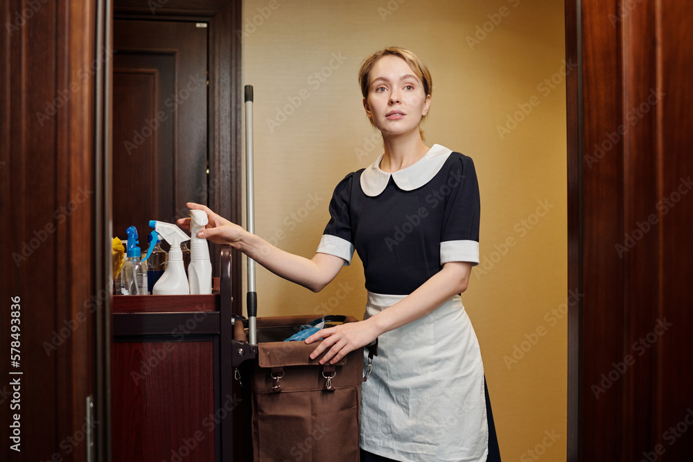 Young pretty room maid in uniform taking bottle with sprayer from cart ...
