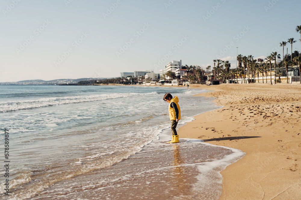 Boy in yellow rubber boots playing with water and sand at the beach ...