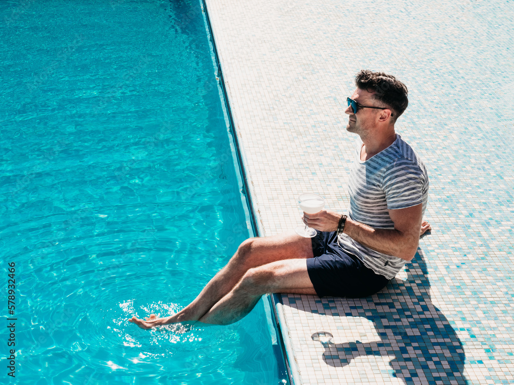 Handsome man with a glass of drink sits near the swimming pool of a ...