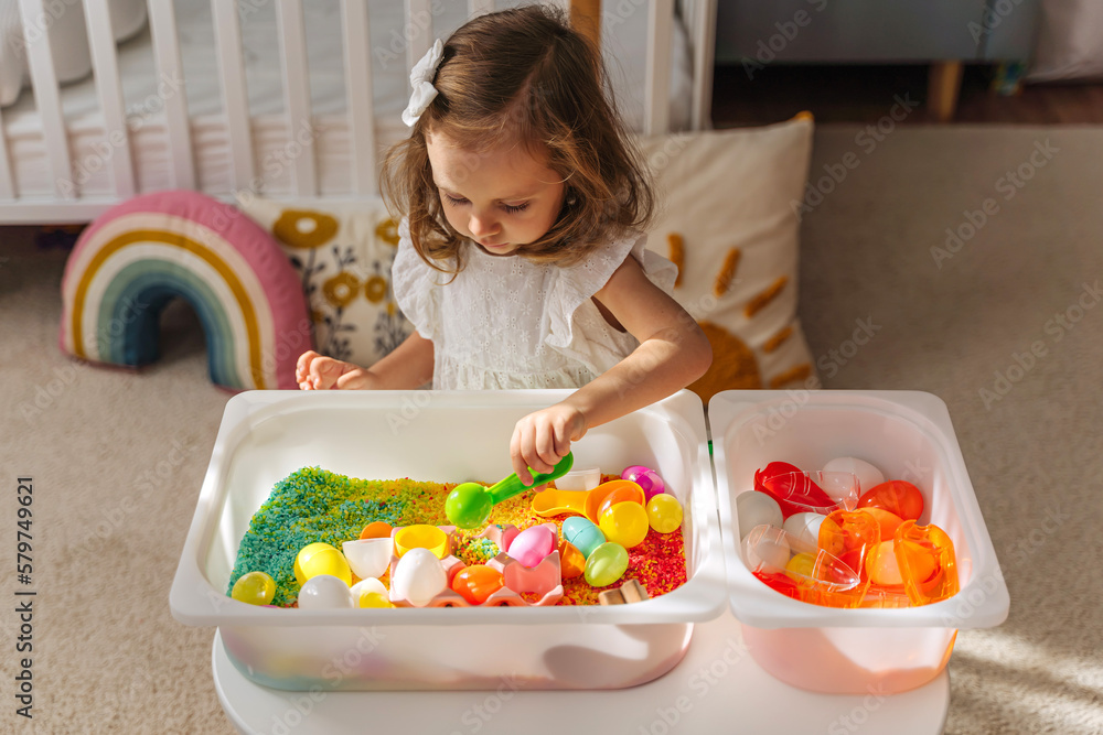 A little girl playing with colored rice and Easter eggs in sensory bin ...