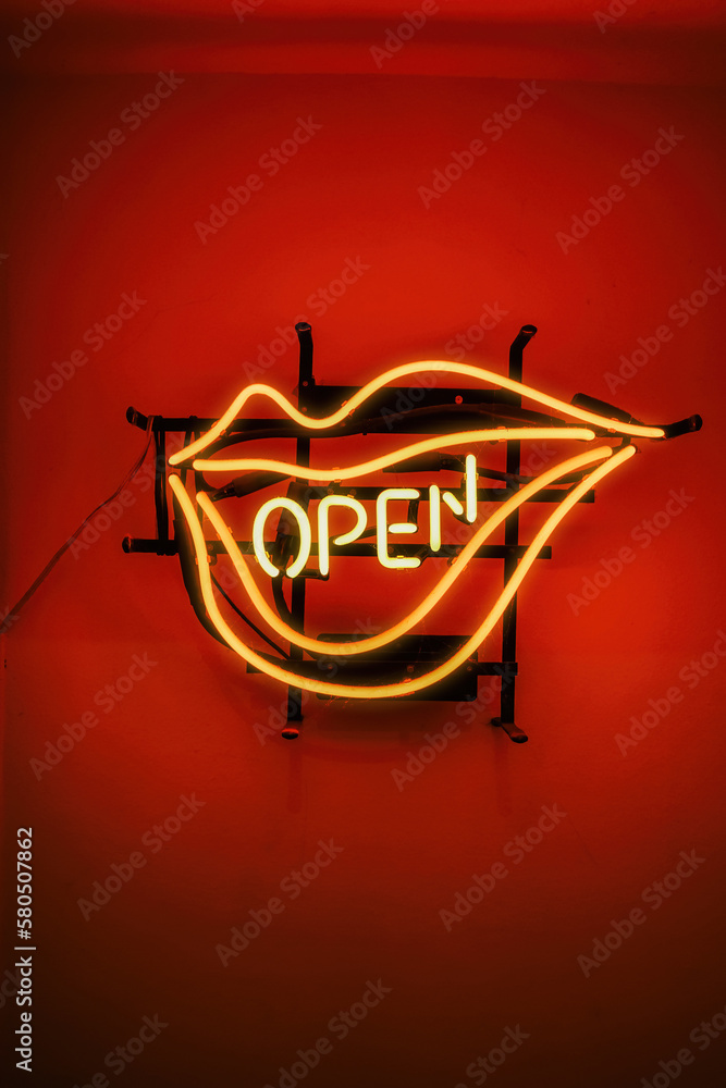 Orange neon sign of an open mouth glows against a red wall in a bar. In ...