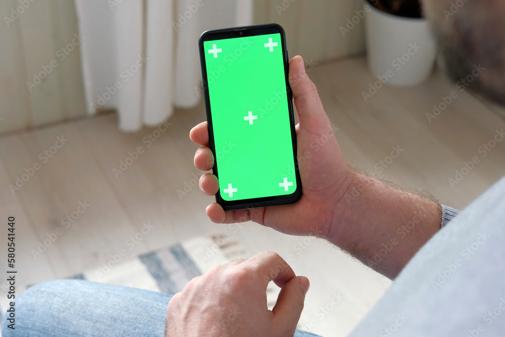 Young man sitting at home holding smartphone green mock-up screen in ...
