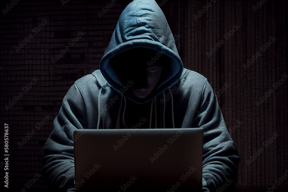 A person in a hoodie typing on a laptop while sitting in a darkened ...