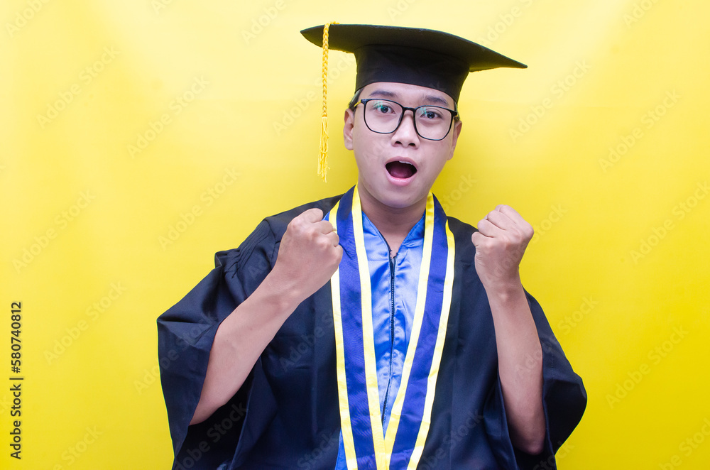 A young Asian man celebrates his graduation by clenching his fist and ...