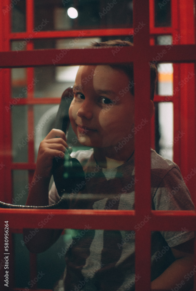 Child boy in a red phone booth smiles and looks at the camera through ...