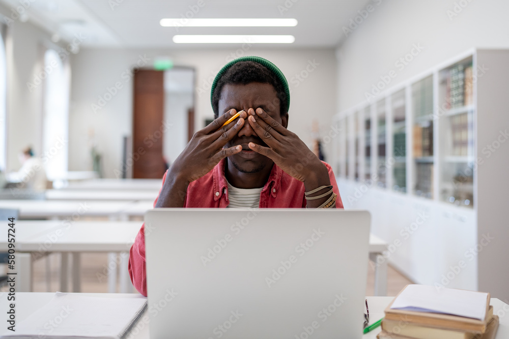 Exhausted African American student man sit at library desk with laptop ...