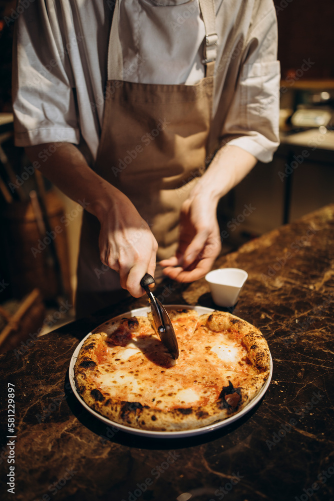 cut the pizza with a round knife into pieces. a man in an apron cuts a ...