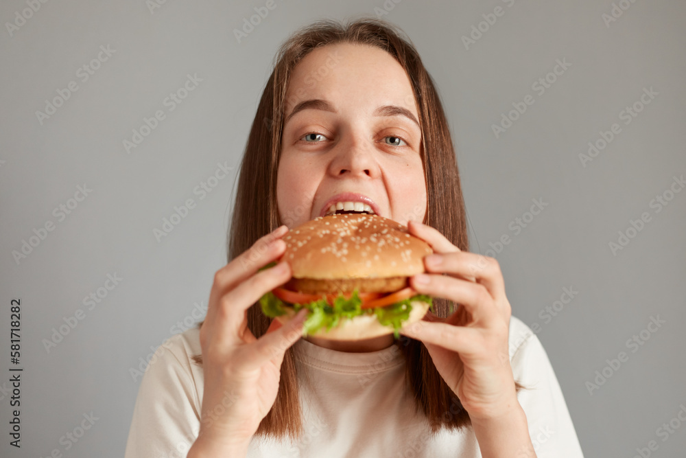 Indoor shot of hungry woman with brown hair biting cheeseburger with ...