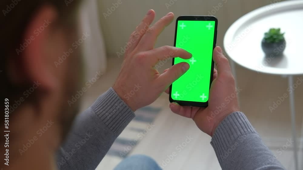 Young man sitting at home holding smartphone green mock-up screen in hand. Male person using ...