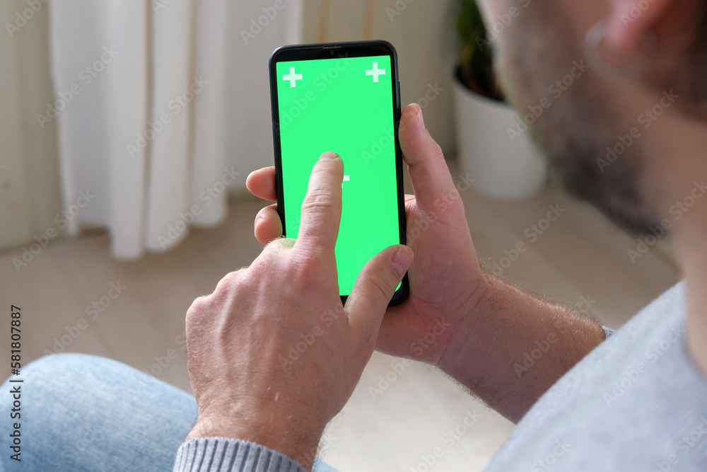 Young man sitting at home holding smartphone green mock-up screen in hand. Male person using ...