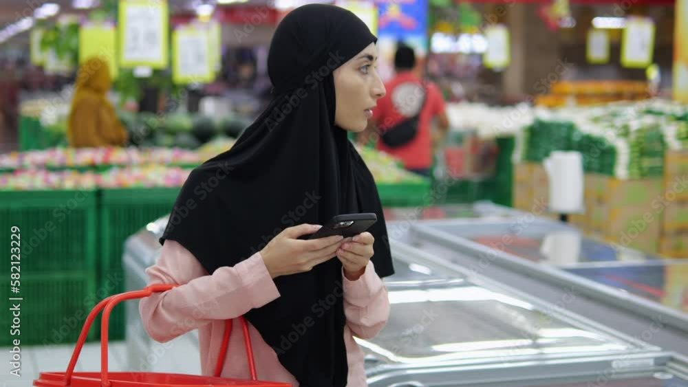 Muslim woman in hijab stands in supermarket with red shopping basket ...