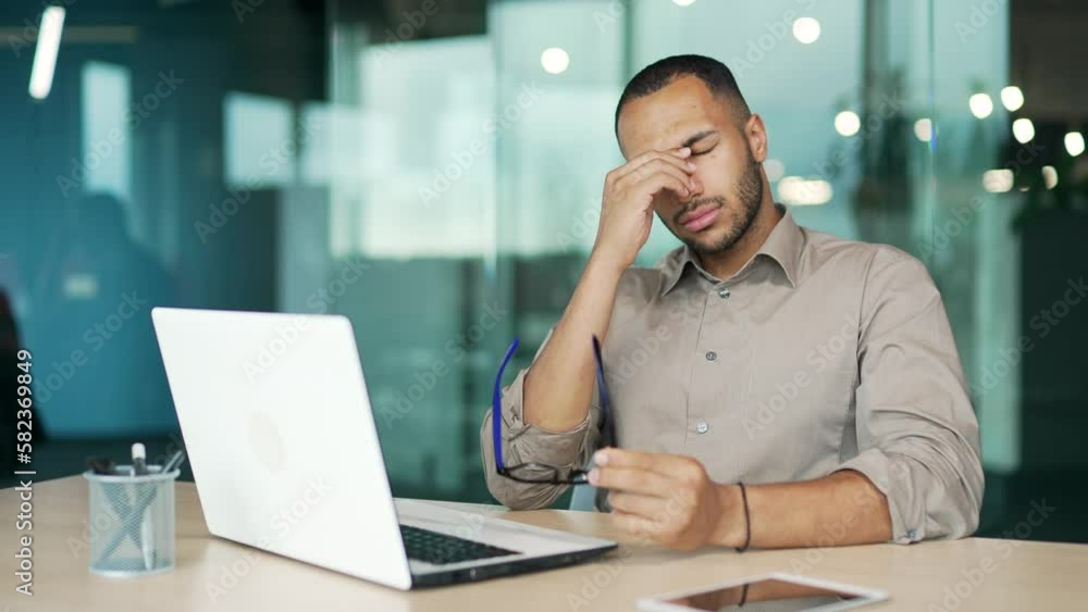 Young adult man tired at work using laptop while sitting at workplace ...