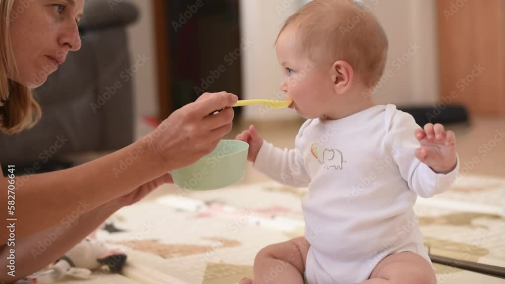 Mother gives food of baby in white bodysuit. Child eating first food ...
