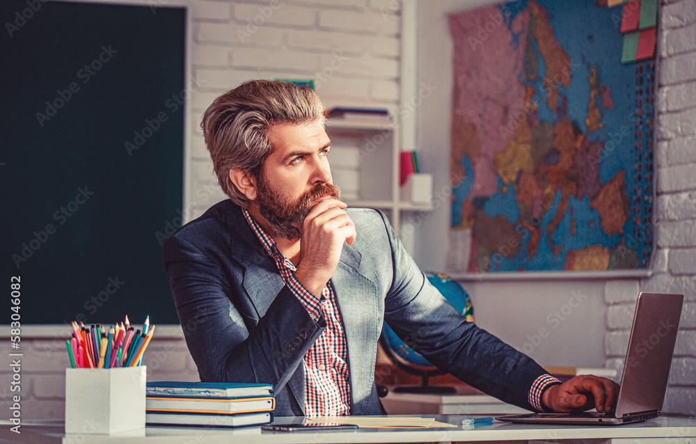 Young male teacher with laptop in class sits at the table the ...