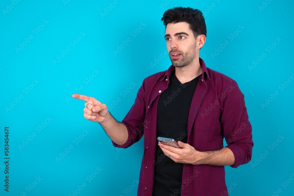 Stunned Young man standing over blue studio background points sideways ...