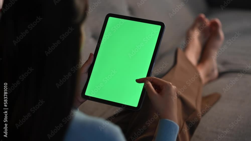 Rear view of young woman relaxing on couch, scrolling at green screen ...