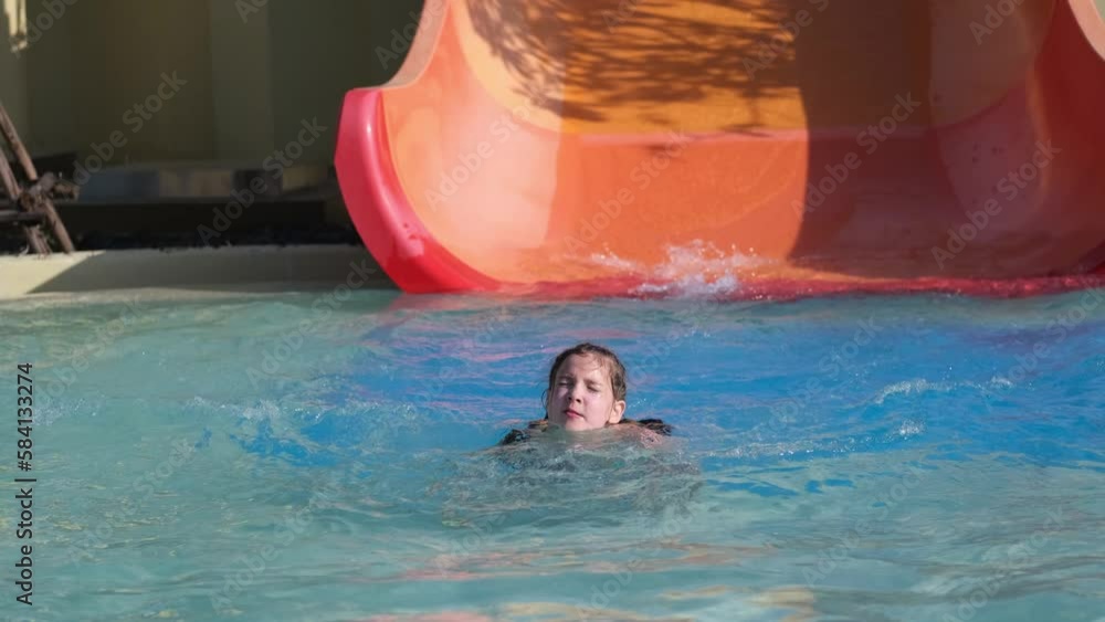 a little girl in a purple swimsuit rolls down a multicolored slide into ...