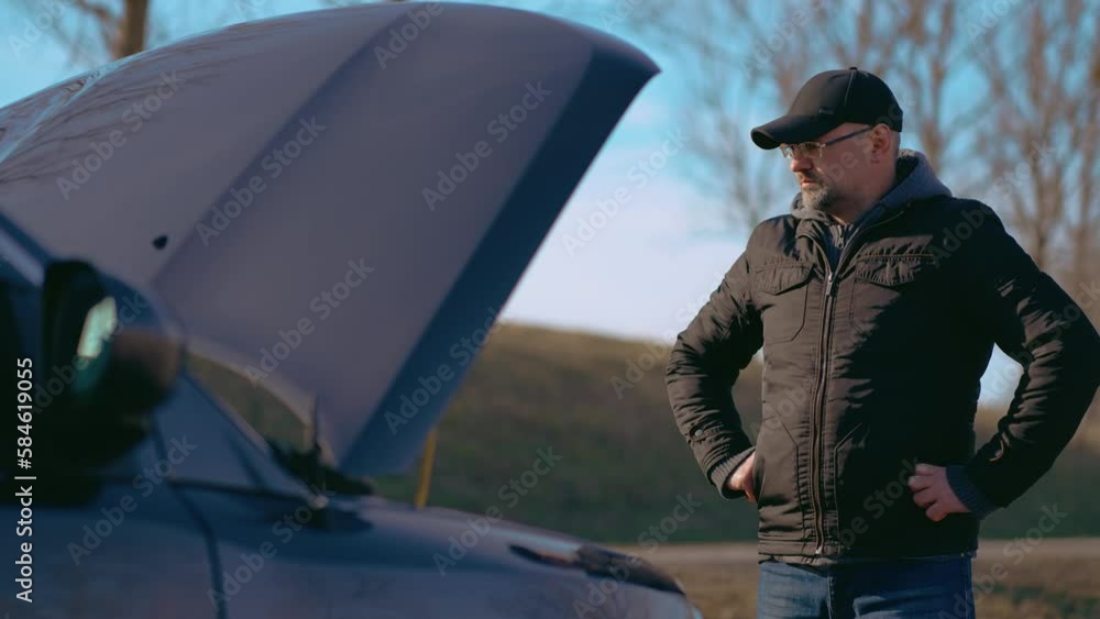 Frustrated man standing next to his broken car with open hood, looking ...